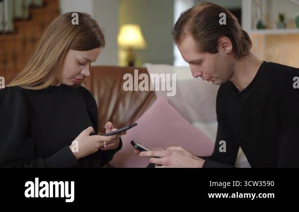 A young man and woman sit together on a sofa, each focused on their ...