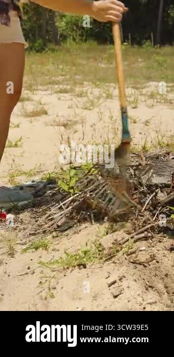 Vertical video. Woman raking pile of dry leaves and debris on sandy ...