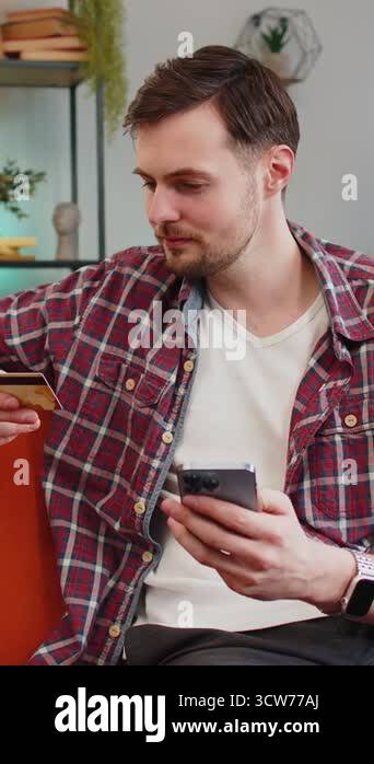 Sad young man guy in plaid shirt sitting on sofa holding smartphone and ...