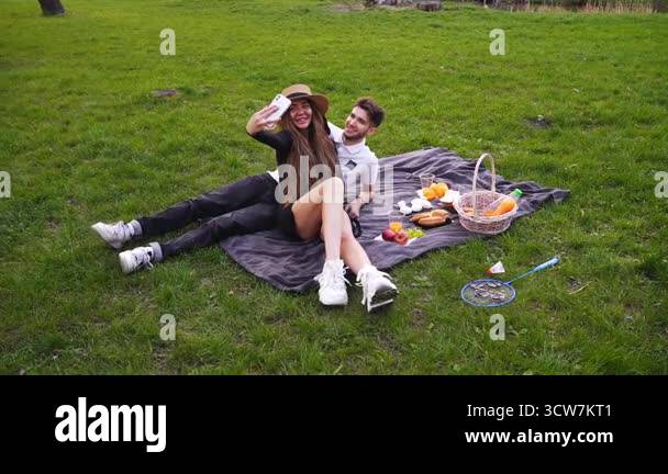 young couple in love lies on blanket in park and takes selfie on smartphone, man and woman relax ...