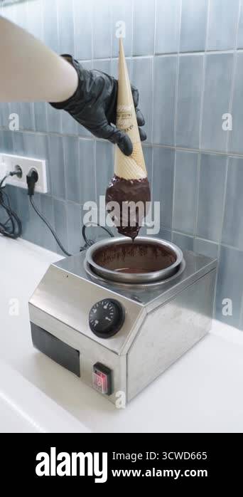 Pastry chef in black gloves preparing ice cream cone, dipping it into a chocolate warmer. Slow ...