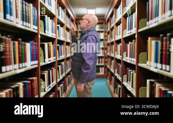 A focused man stands in a library aisle, examining books on the shelves ...