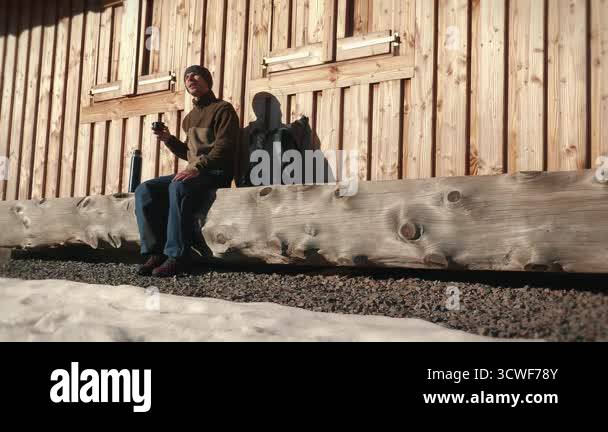 Man hiker sitting on bench near forest cabin, drinking tea and enjoying ...