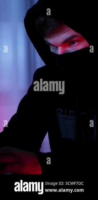 Male in black mask and hood looking at laptop screen and typing on ...
