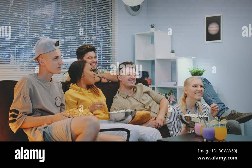 Multi-ethnic young people watching sports on TV while eating snacks at ...