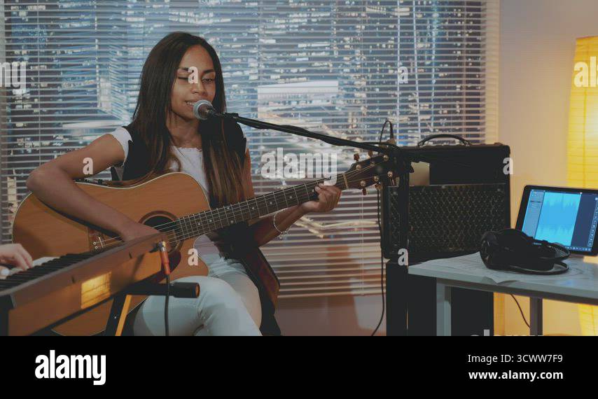 Mixed-race young lady playing guitar and singing into microphone during band rehearsal in home ...