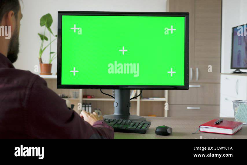 Side view of man sitting in front of computer with big green screen ...