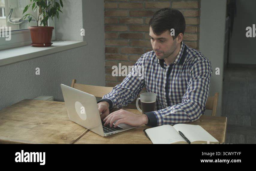 A man in a plaid shirt, stares at the monitor, takes a sip of coffee ...