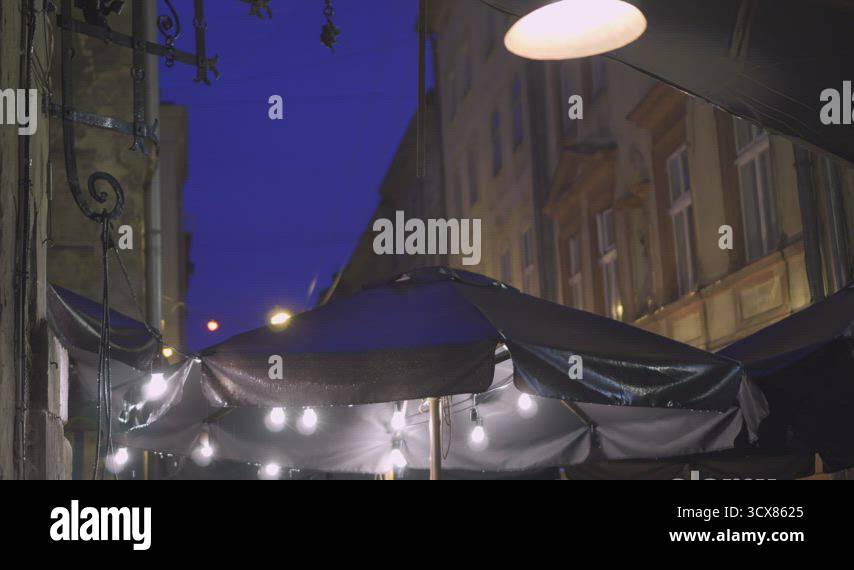 Outdoor street cafe at night in heavy rain. Raindrops falling down the canopy. Lanterns hanging ...