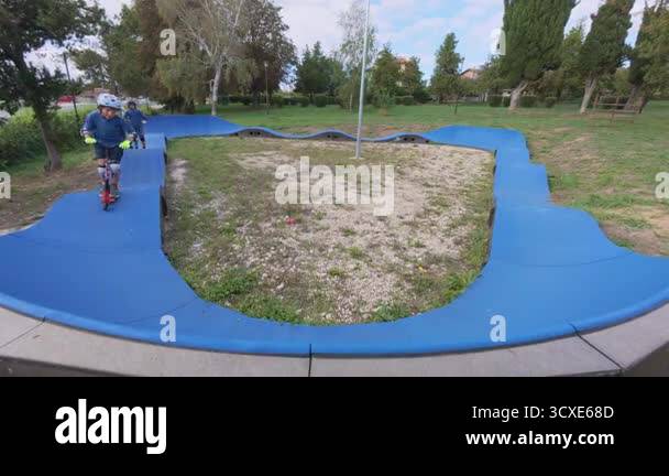 Two children riding scooters on a blue pump track in a park, wearing ...