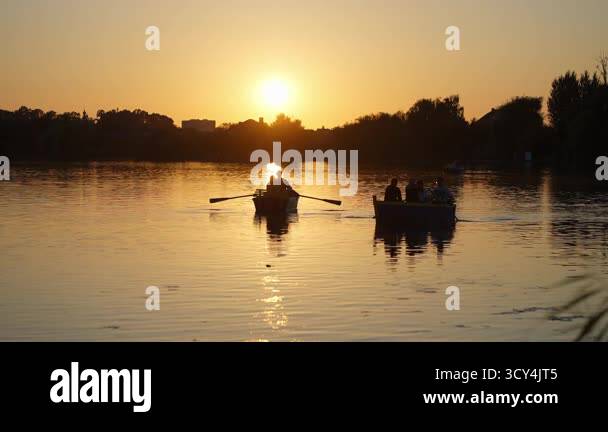Boats on lake during golden sunset, people rowing on calm water with ...