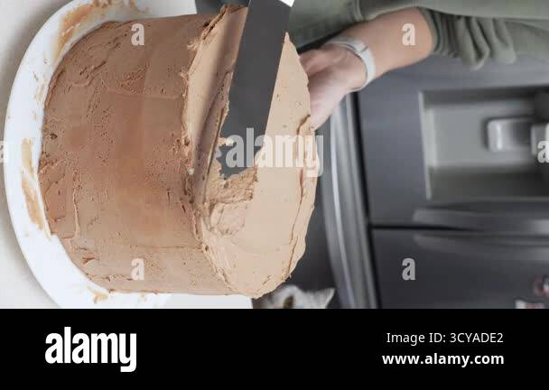 A young woman is frosting a chocolate cake using a spatula. The cake is ...