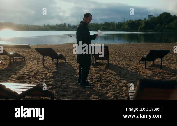 A somber man stands alone on a quiet beach, reading a handwritten ...