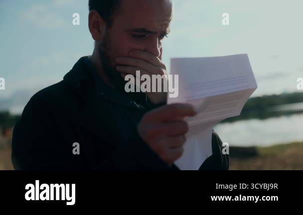 A distressed man stands alone outdoors reading a letter. His expression ...