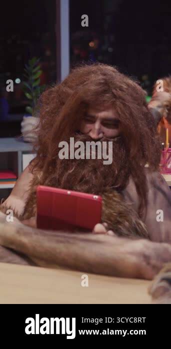 Bearded prehistoric caveman in fur clothes sitting on office desk using ...
