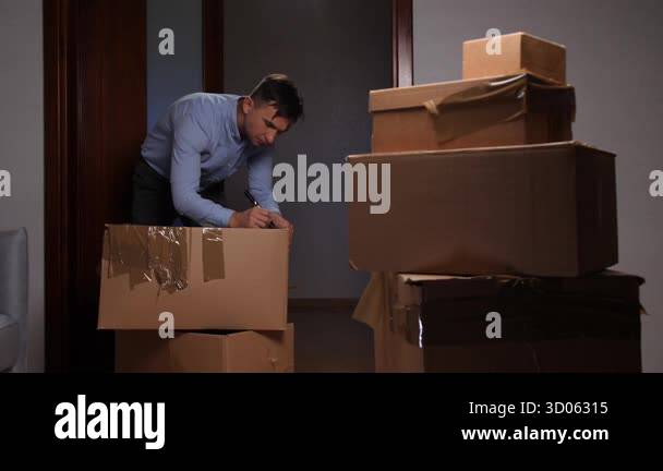 Man labeling a cardboard box with a marker amid packed boxes, preparing ...