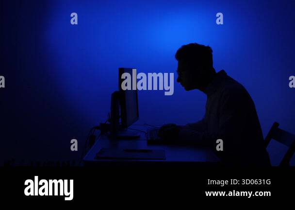 Man in silhouette sitting at a desk, intensely focused on a computer ...