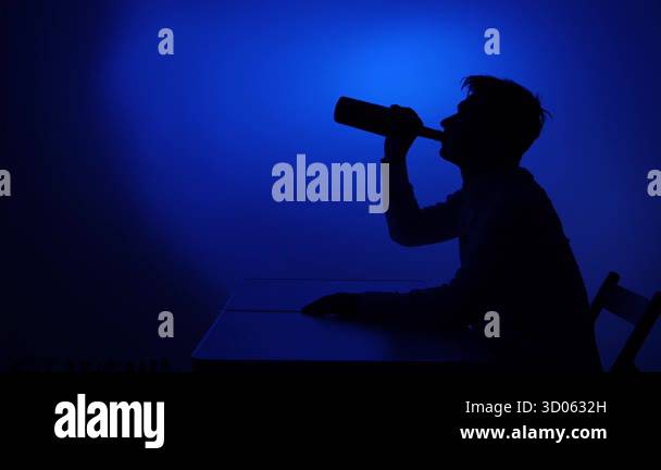 Silhouette of a man sitting alone at a table drinking from a bottle ...