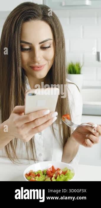 A woman sits in a bright kitchen, happily eating a salad while looking ...