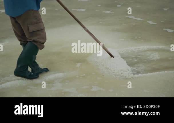 Laborer in rubber boots gathers wet salt with wooden shovel in ...
