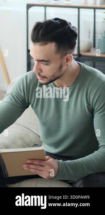 A thoughtful young adult man sits on a sofa at home, thinking and ...