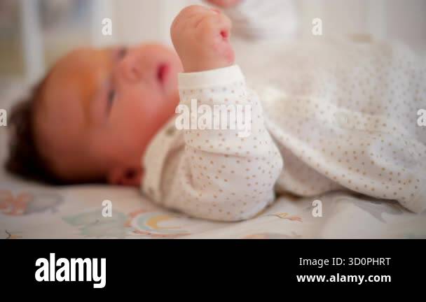 A newborn baby in soft pajamas moves gently in a crib, stretching tiny ...
