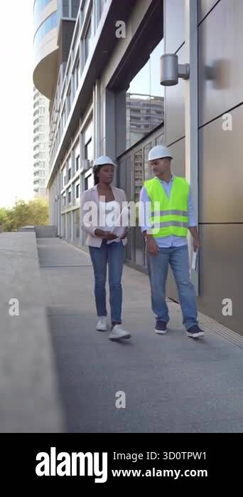 Two diverse architects in safety vest and helmets walking and talking ...