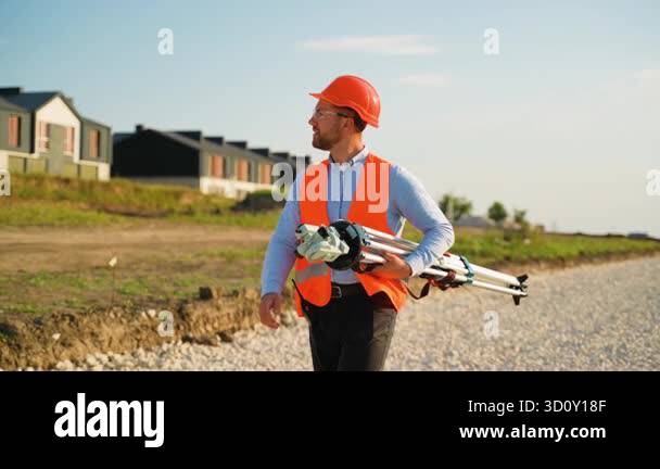 Caucasian land surveyor wearing a hard hat and safety vest walking on a ...