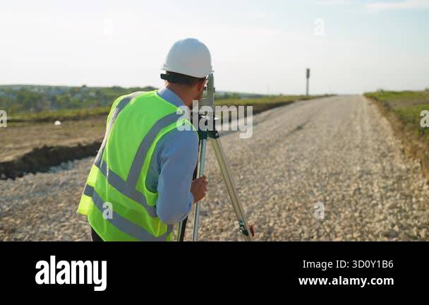 Male surveyor wearing a hard hat and safety vest performing geodetic ...