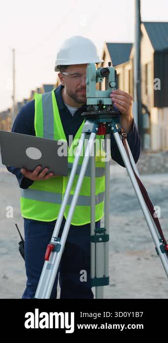 Professional male land surveyor in a hard hat and safety vest using a ...