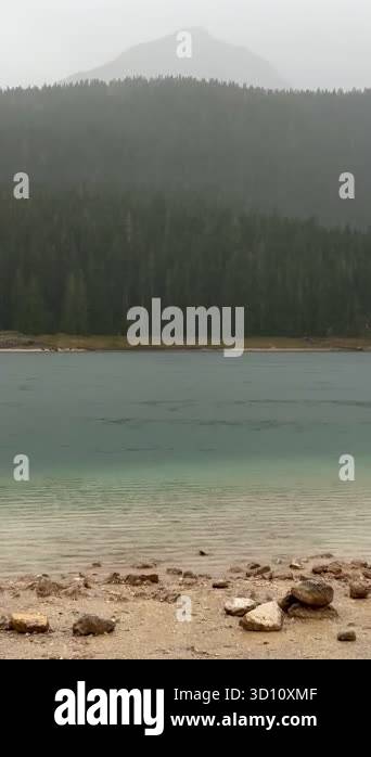 Cinematic view of rainfall over calm turquoise lake with dense conifer ...