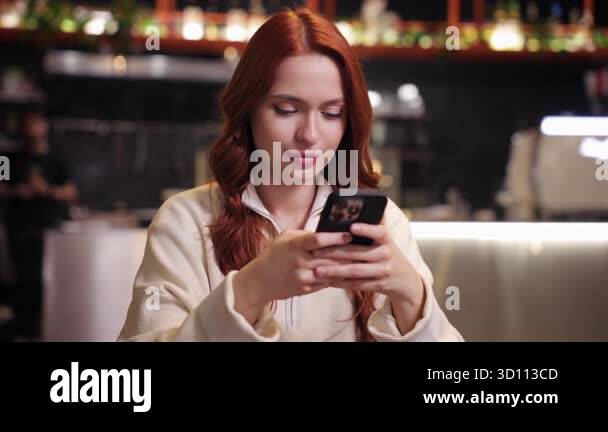 Focused red-haired girl gazing at smartphone while sitting in modern ...