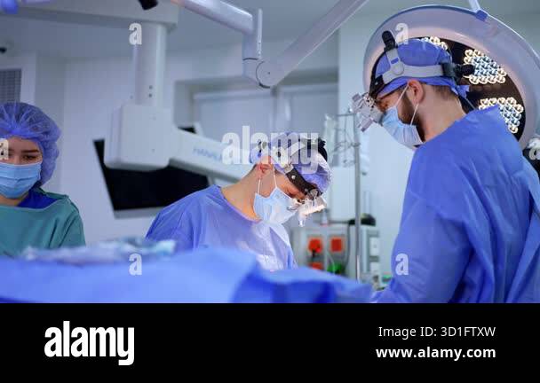 Two male neurosurgeons wearing surgical loupes stand at the round portable lamp. Scrub nurse ...