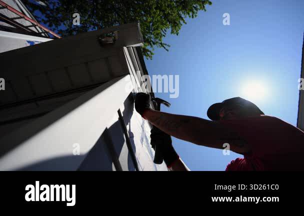Worker hummers a nail into the plastic panel siding on the outside wall of the residential house ...