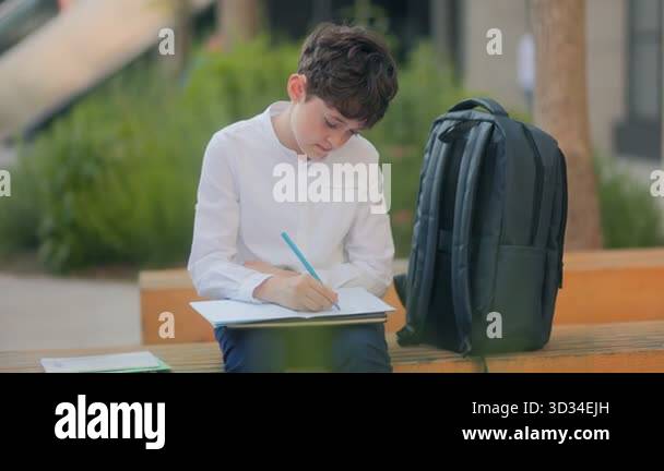 School-age boy sits on a bench doing his homework outside with a black ...