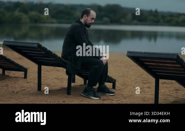 Bald man sits on a bench by the lake, hunched over in deep sadness. The ...