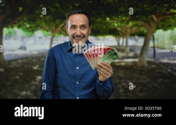 Smiling man in park holds israeli shekel banknotes showcasing vibrant ...