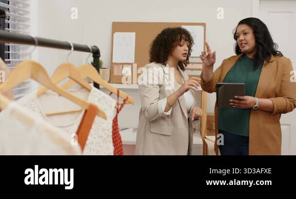 Female co-workers reviewing tablet and browsing red garment on rack for ...