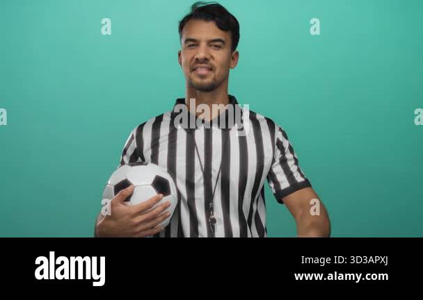 Young hispanic male referee holding soccer ball smiling pointing ...