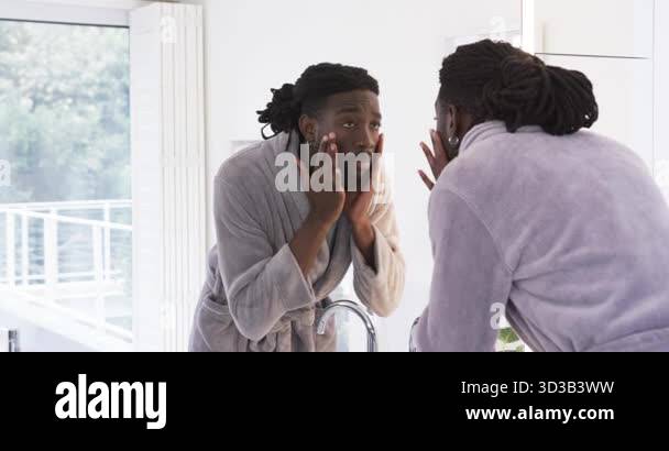 African American man in robe checking mirror spreading cream patting ...