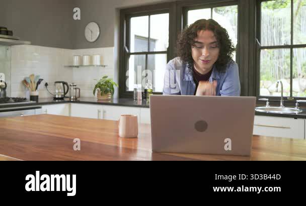Non-binary adult scrolling on laptop and sipping coffee at home ...