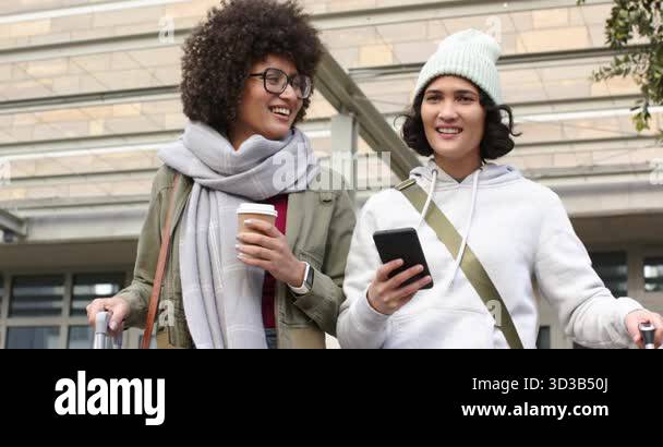 Diverse female friends walking outside building, scrolling phone ...