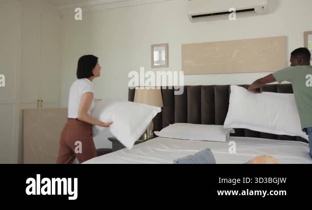 Diverse couple entering bedroom at home, unpacking box, placing duvet ...
