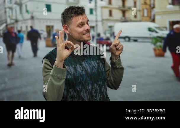 Young man gesturing okay and pointing sideways with a smile in an urban ...