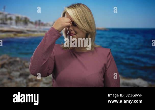 Blonde woman in rose blouse pinches nose with closed eyes while ...