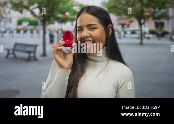 Woman smiling holding a red engagement ring box outdoors on a street ...