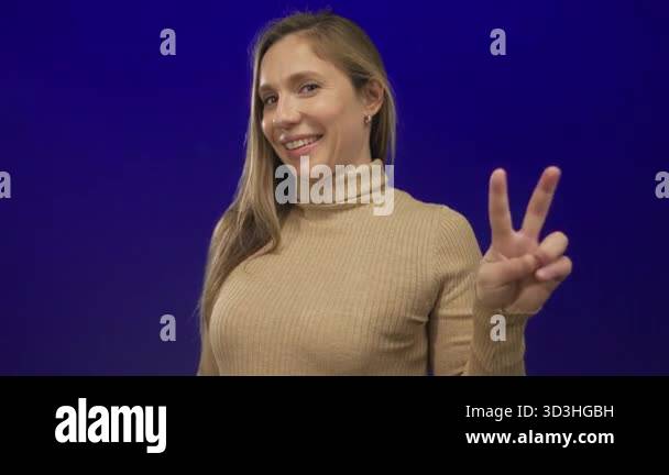 Woman smiling and making a peace sign in front of a blue background ...