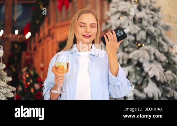 Joyful woman smiles while using smartphone, holding glass of wine ...
