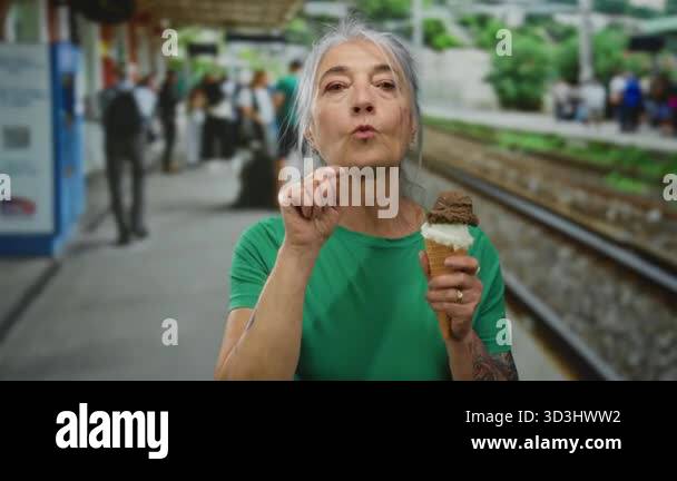 Senior woman with grey hair at a train station enjoys an ice cream cone ...
