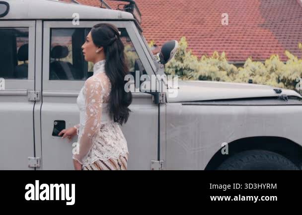 Elegant woman in a white dress opening the door of a vintage car during ...